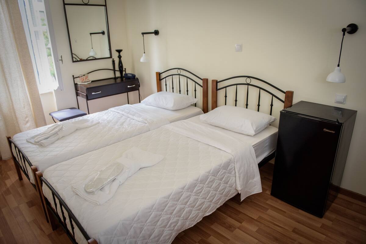 Uma cama de casal com roupa de cama branca, uma mesinha de cabeceira pequena, uma luminária e um frigorífico.