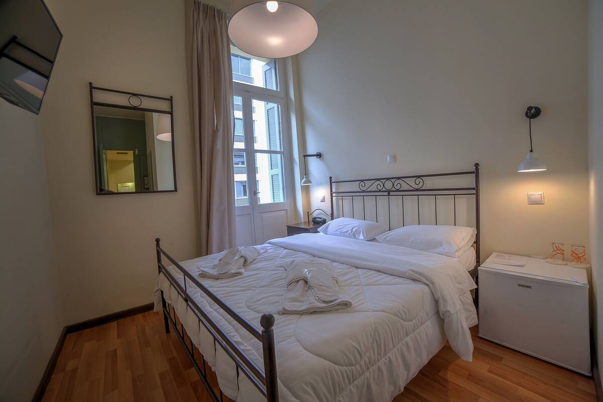 Cama bem arrumada com toalhas, mesa de cabeceira com luminária, pequeno refrigerador e janela com cortinas.