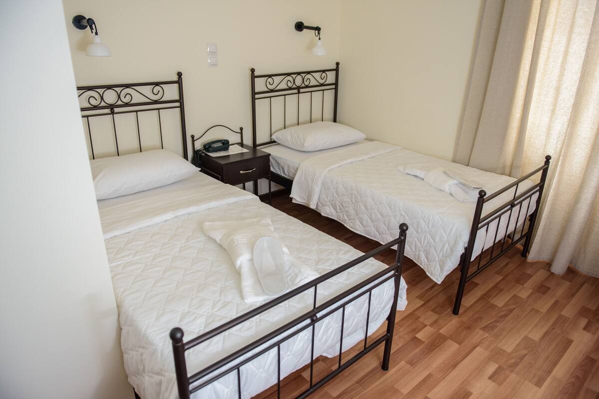 Quarto com duas camas de solteiro, cada uma com colchão e almofadas brancas acolchoadas. Há duas mesas de cabeceira com luminárias, e o chão é de madeira.