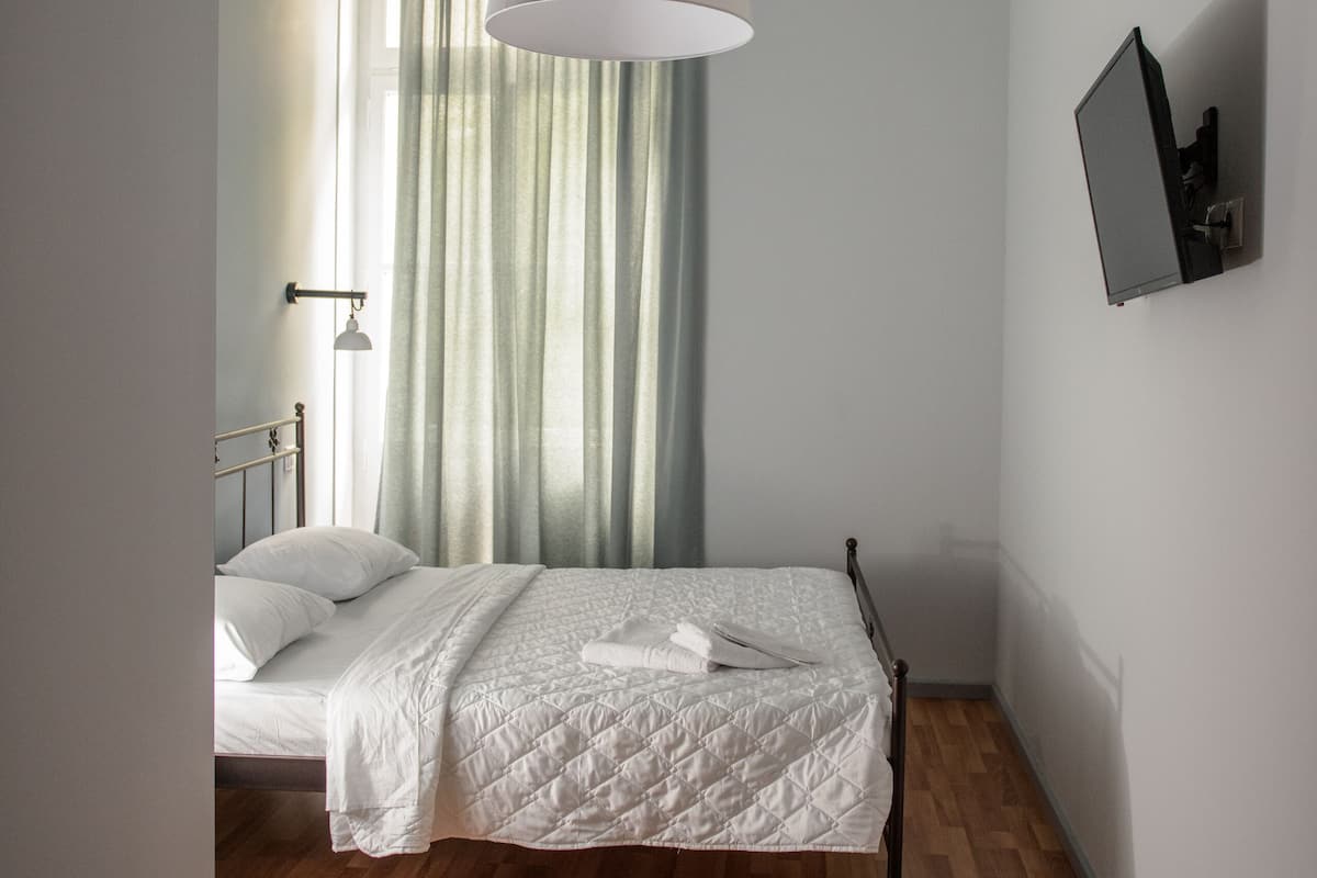 Quarto com cama, piso de madeira, luminária pendente, janela com cortinas e televisão na parede.
