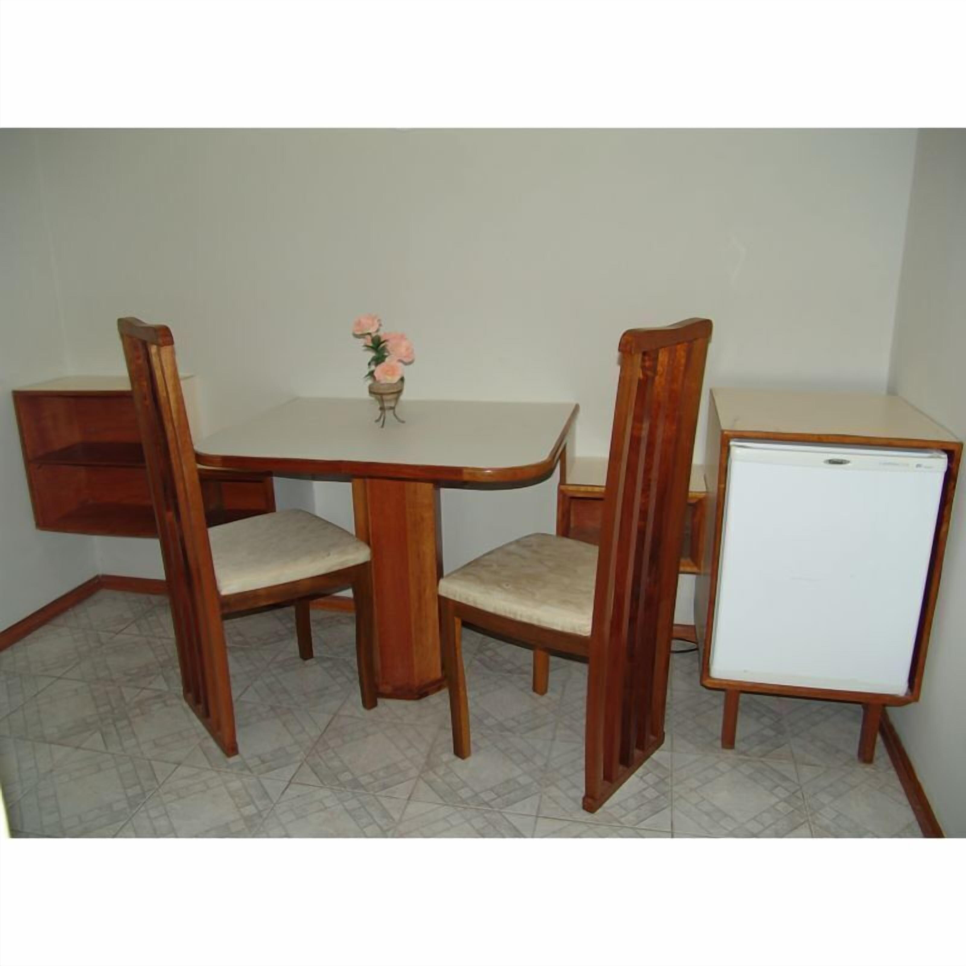 Uma pequena área de refeições com mesa e cadeiras de madeira, um vaso com flores e uma geladeira branca.