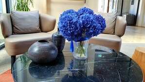 Um hall moderno com uma mesa de centro de mármore preto, dois sofás bege, uma planta em vaso e um arranjo de flores azuis.