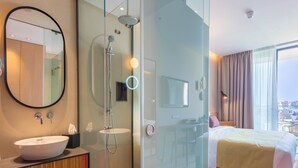 Quarto de hotel moderno com uma cama grande, banheiro com box de vidro, espelho redondo e vista para a cidade.
