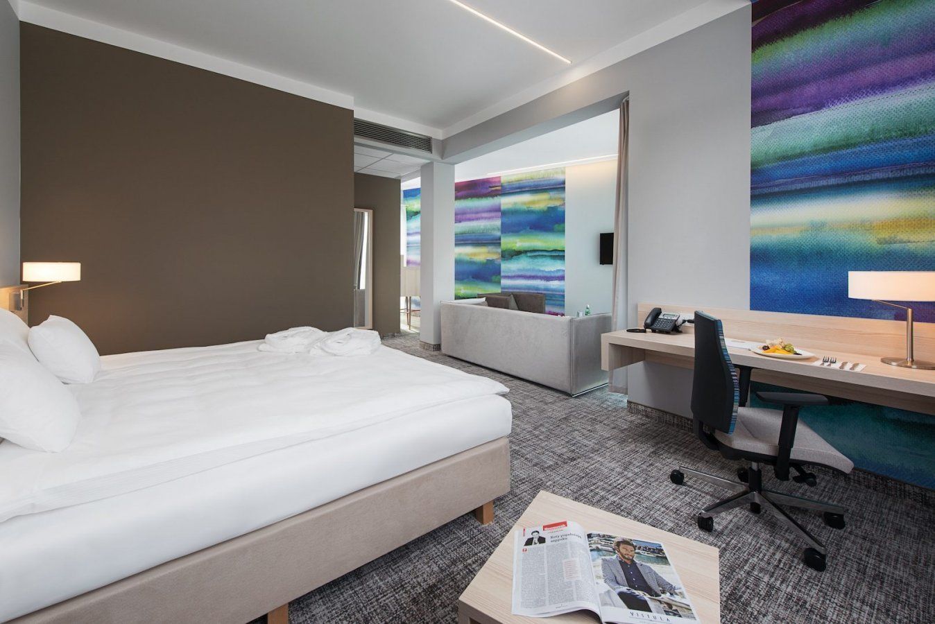 Quarto de hotel com cama, escrivaninha, cadeira, abajur e uma revista no chão.