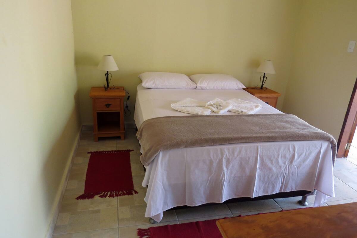 Uma cama bem arrumada com lençóis brancos e um cobertor marrom, duas luminárias de cabeceira e um tapete vermelho sobre um piso de azulejos.