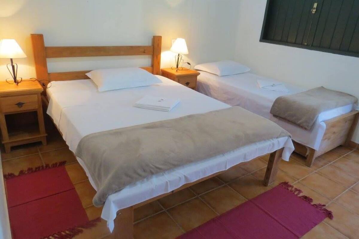 Quarto de hotel com duas camas de solteiro, cada uma com uma mesa de cabeceira e abajur. Há uma pequena mesa com uma cadeira e uma janela com persianas.
