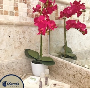 Um banheiro com pia branca, espelho e uma planta em vaso com flores rosa.