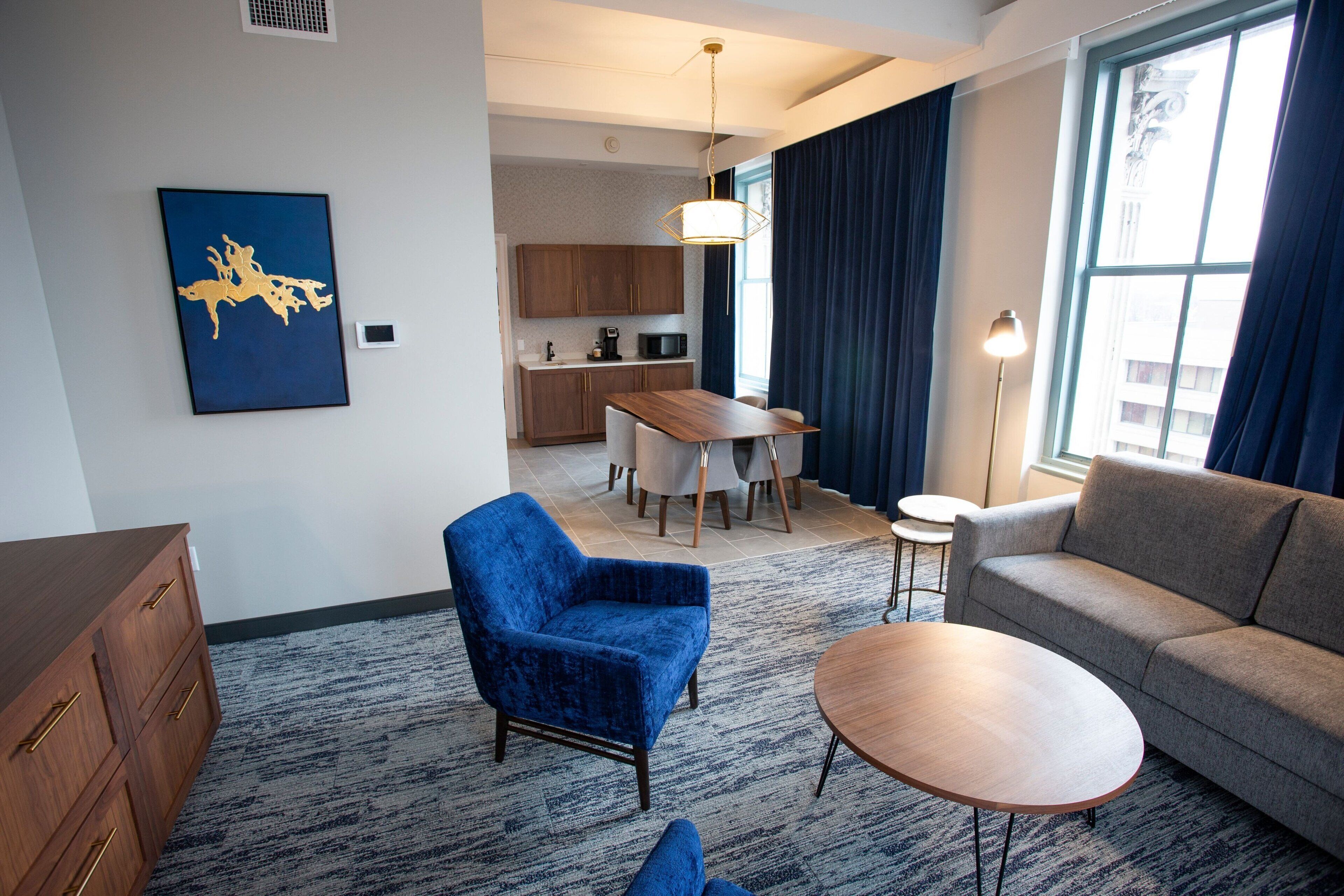 Quarto de hotel moderno com poltrona azul, sofá cinzento, mesa de centro de madeira e área de refeições com cozinha ao fundo.