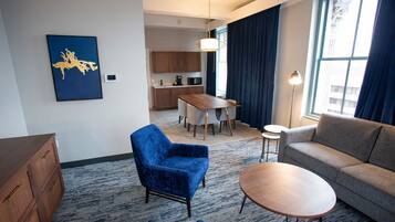 Quarto de hotel moderno com poltrona azul, sofá cinzento, mesa de centro de madeira e área de refeições com cozinha ao fundo.