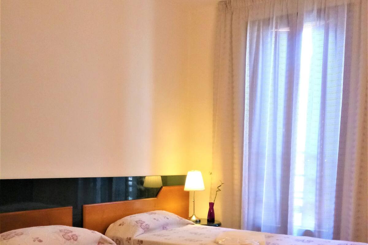 Quarto de hotel com duas camas, uma mesa de cabeceira, um abajur e uma janela com cortinas.