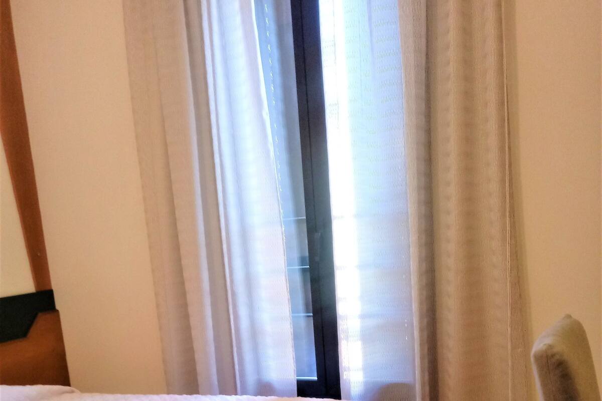 Uma cama bem arrumada com edredom branco e almofadas, cabeceira de madeira, uma cadeira e uma janela com cortinas leves.