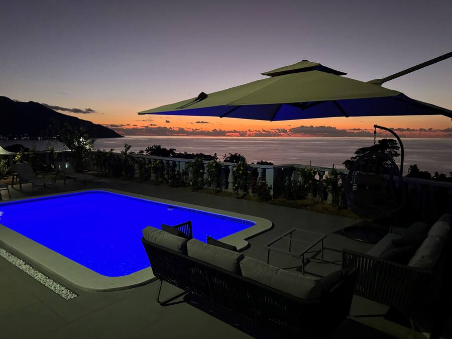 Piscina com vista para o pôr do sol sobre o oceano, cercada por cadeiras de praia e guarda-sóis.