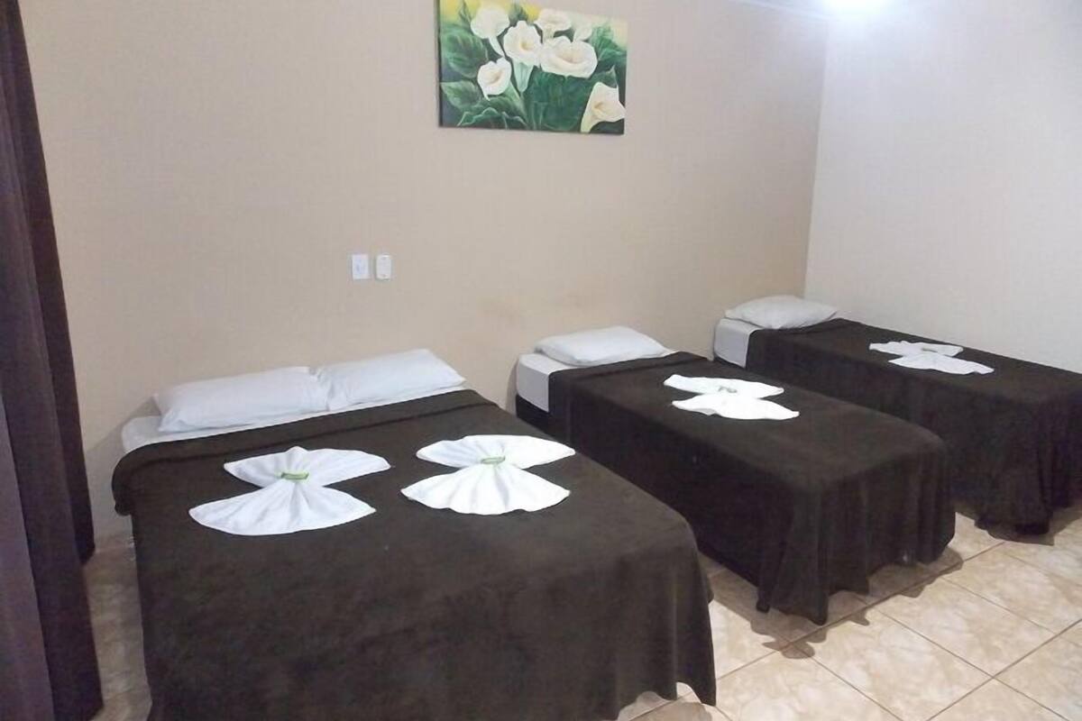 Quarto de hotel com três camas, cada uma com cobertura escura e almofadas brancas, e um quadro de flores na parede.
