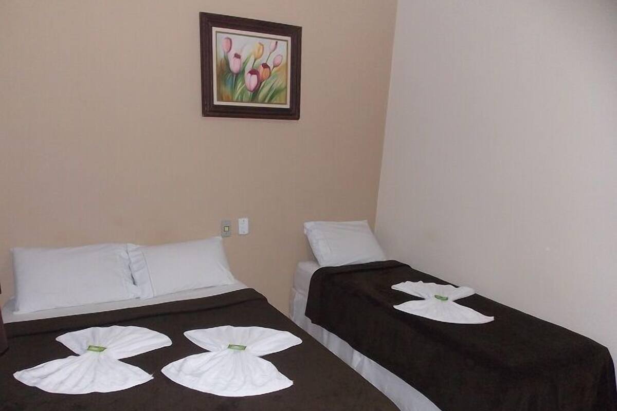Duas camas de solteiro com cobertores marrom-escuros e travesseiros brancos, um quadro com flores na parede, e uma toalha branca dobrada em cada cama.