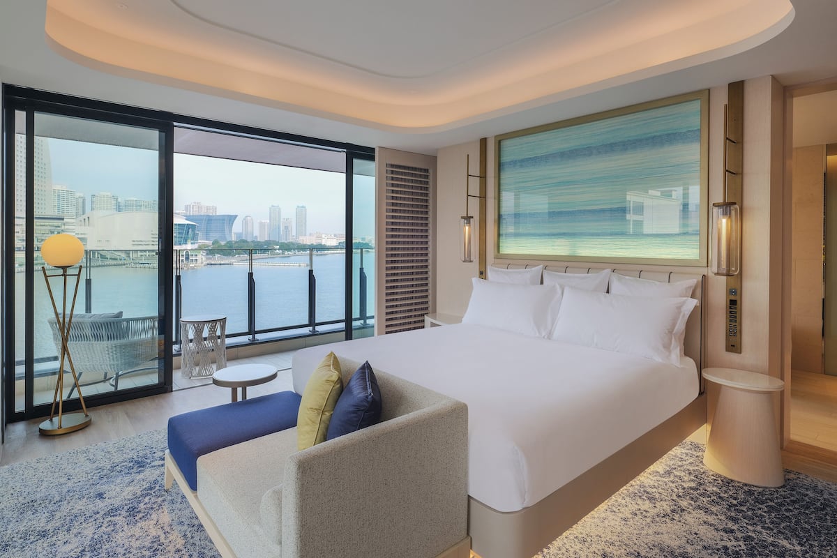 Quarto de hotel moderno com uma cama grande, um sofá, uma mesinha e vista para a cidade e para a água.
