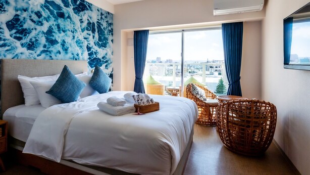 Quarto de hotel com cama, janela ampla com cortinas e vista para a cidade, além de parede com design abstrato azul.