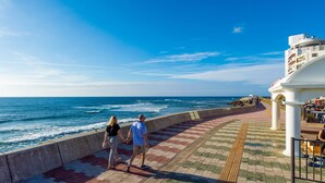 Um calçadão costeiro com piso xadrez, vista para o mar e um edifício branco com varandas.