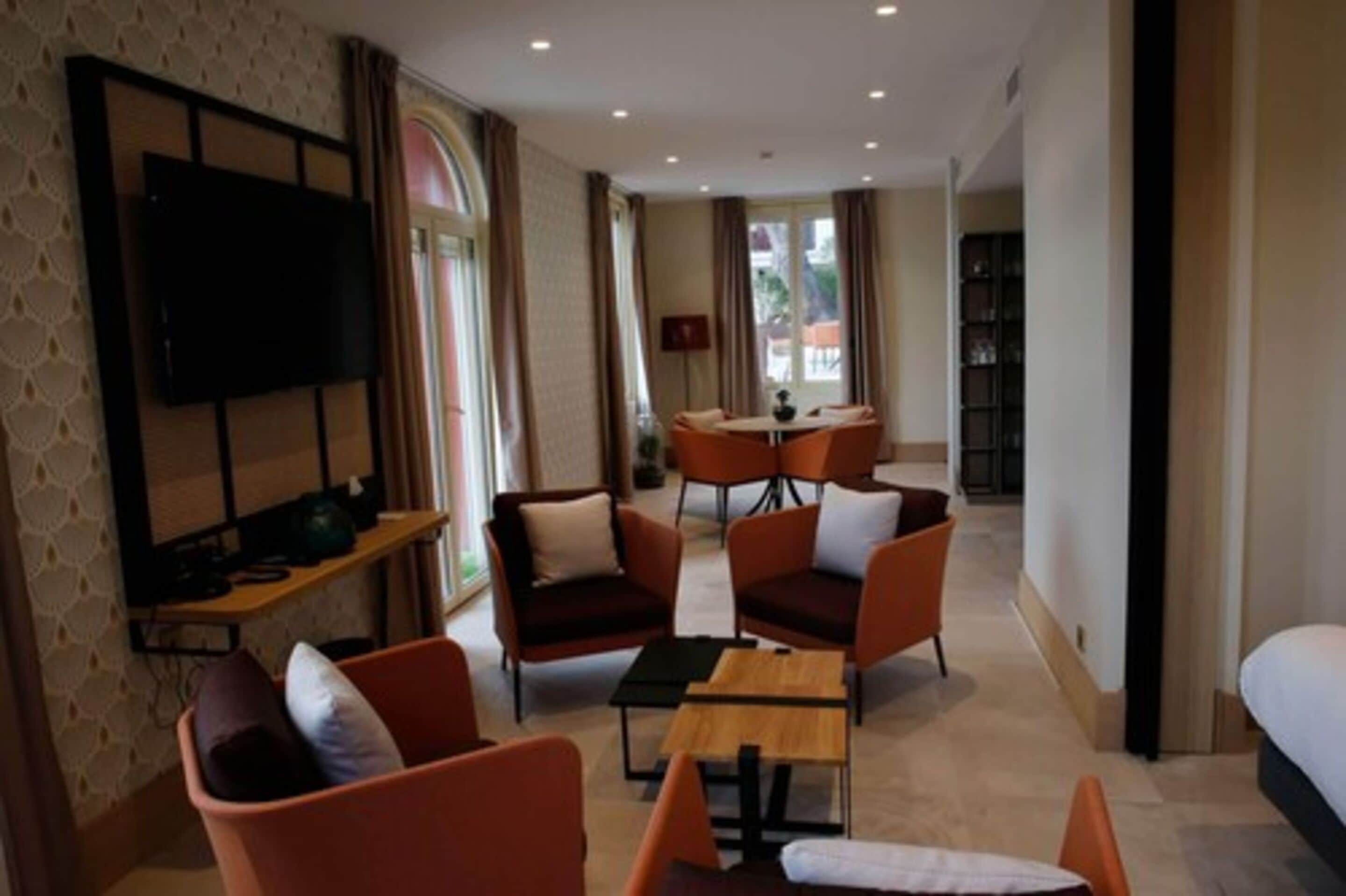 Quarto de hotel moderno com televisão de tela plana, poltronas laranja, uma mesinha e uma cama.