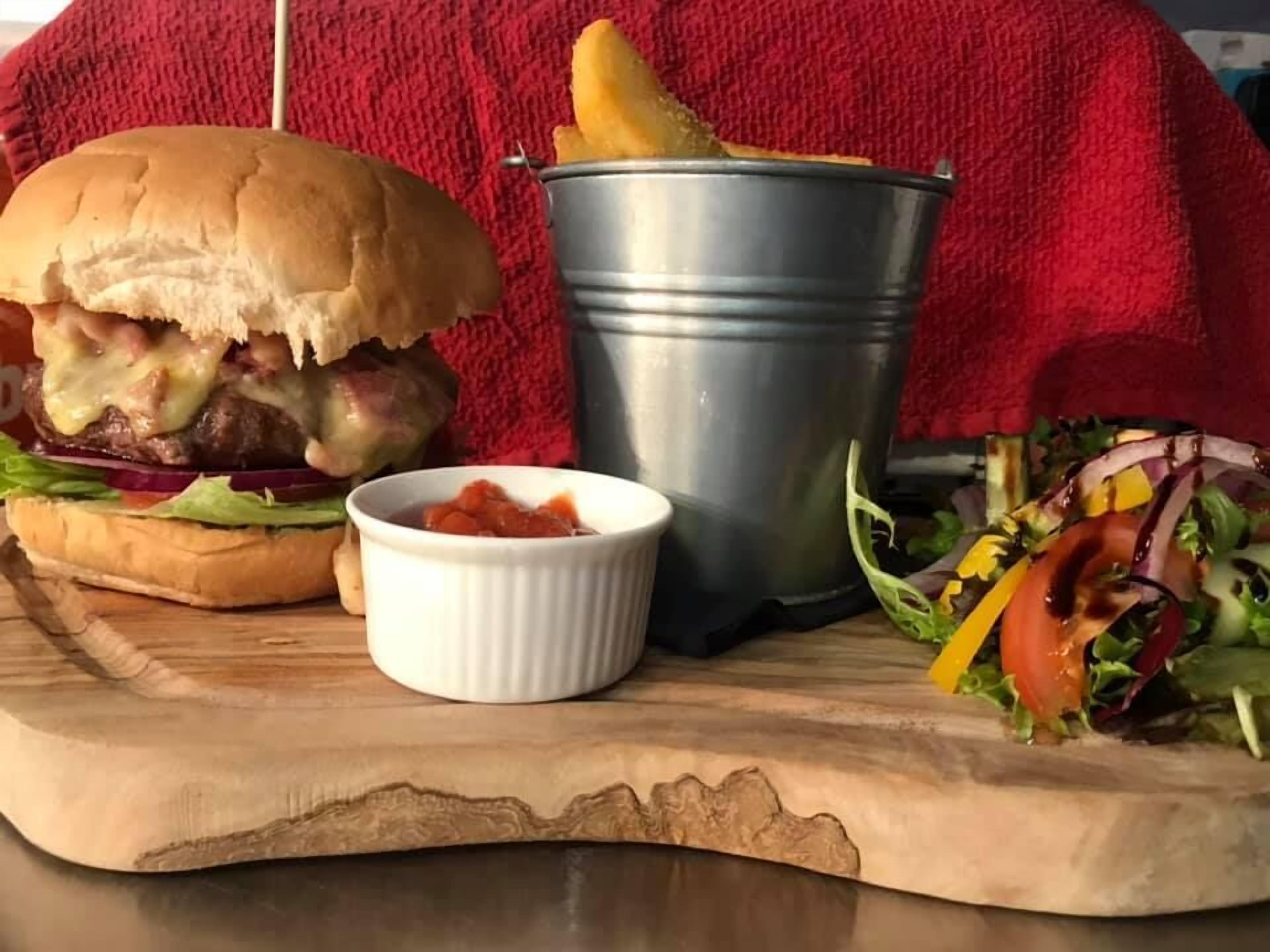 Uma refeição com hambúrguer, batatas fritas, salada e um pequeno recipiente com molho sobre uma tábua de madeira.