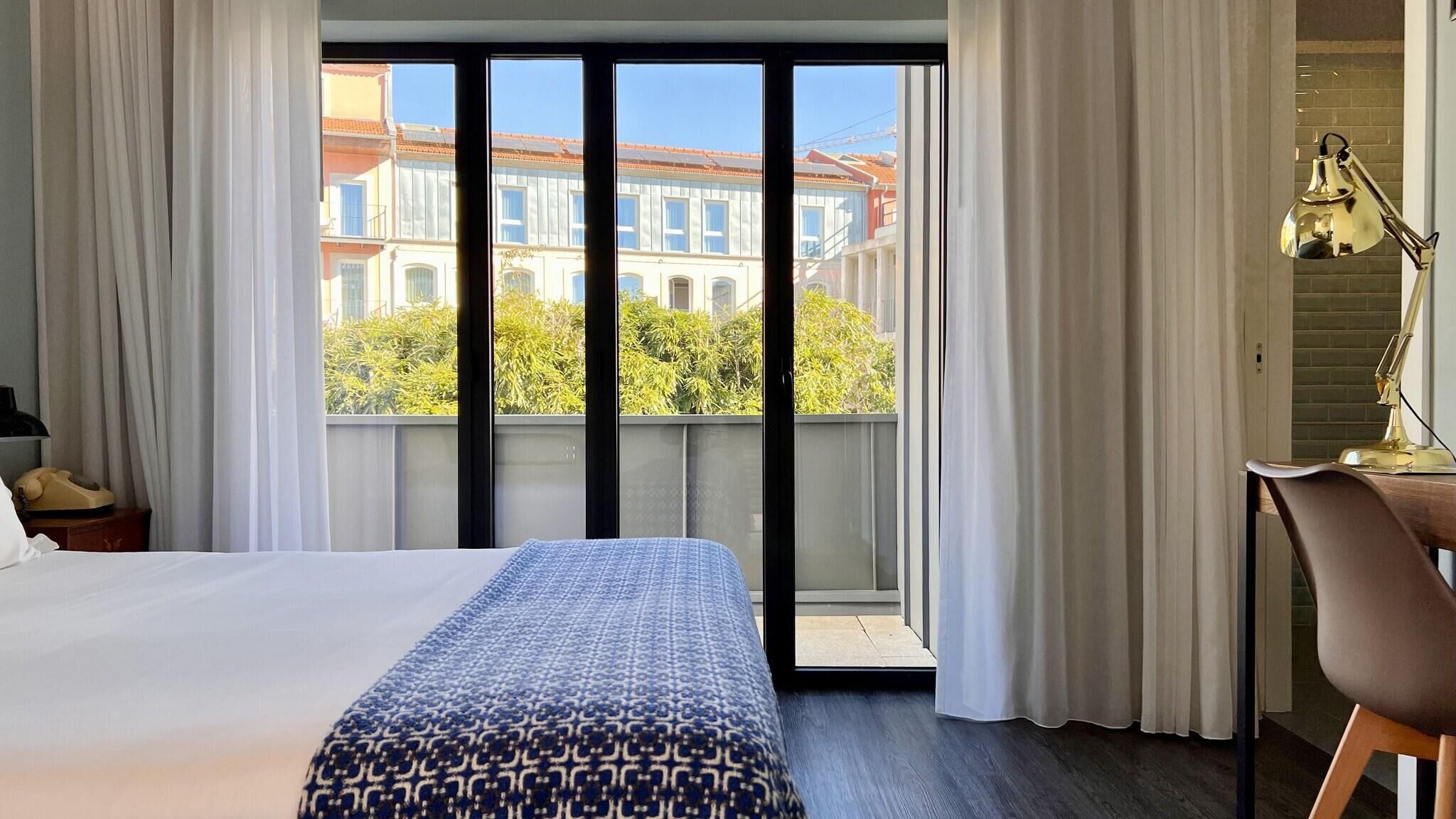 Quarto de hotel com cama, escrivaninha, cadeira e vista para edifícios e árvores através de grandes janelas.