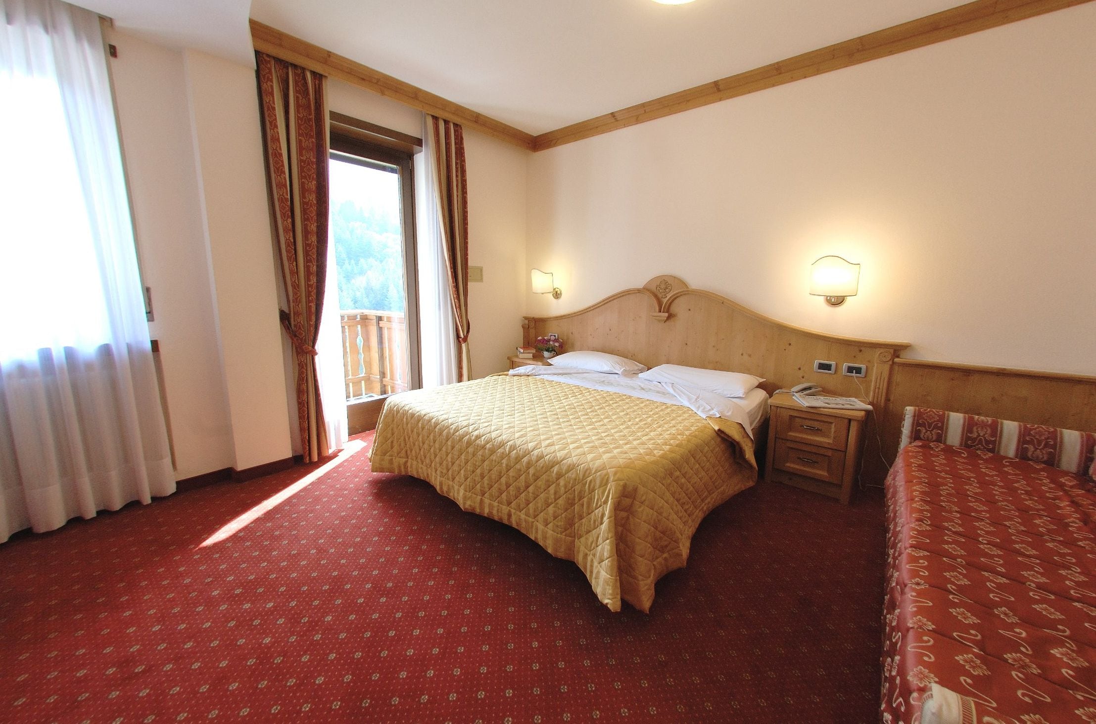 Quarto de hotel com uma cama grande, uma cama menor, uma mesa de cabeceira, uma luminária e uma janela com cortinas.