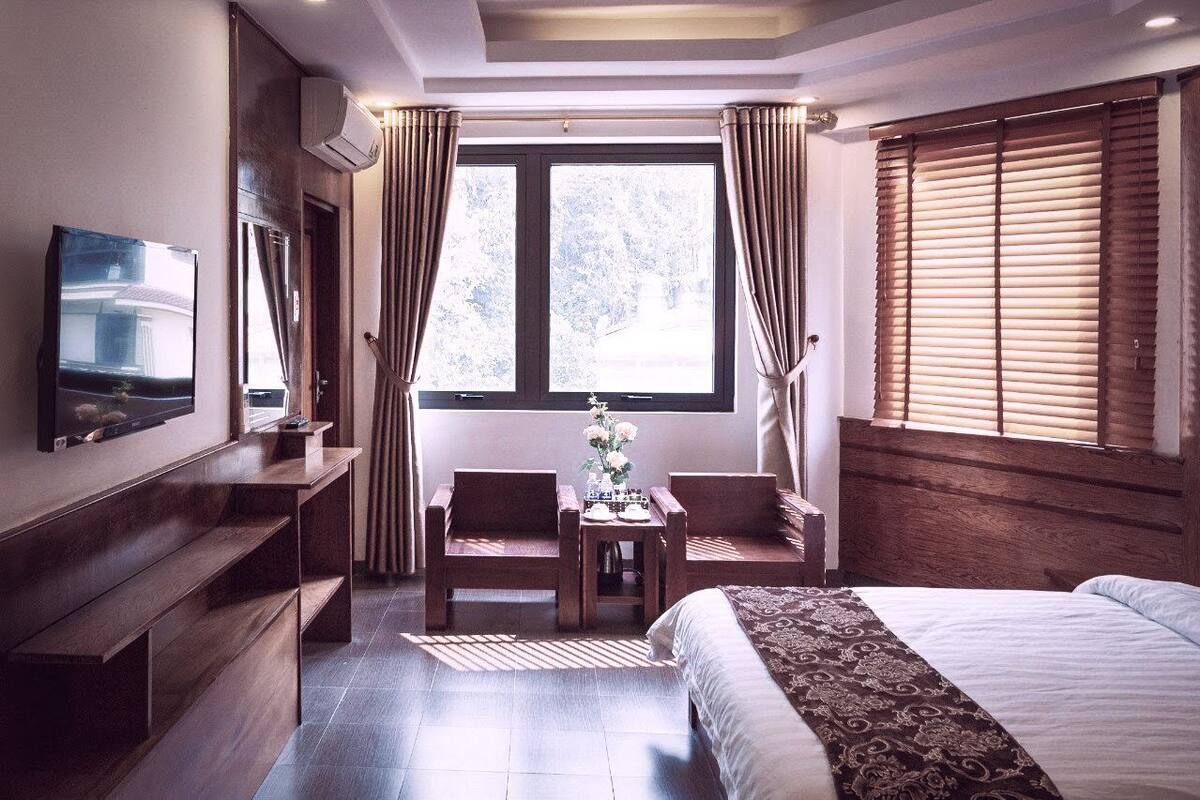 Quarto de hotel com cama, televisão, escrivaninha com cadeiras e janela com cortinas.