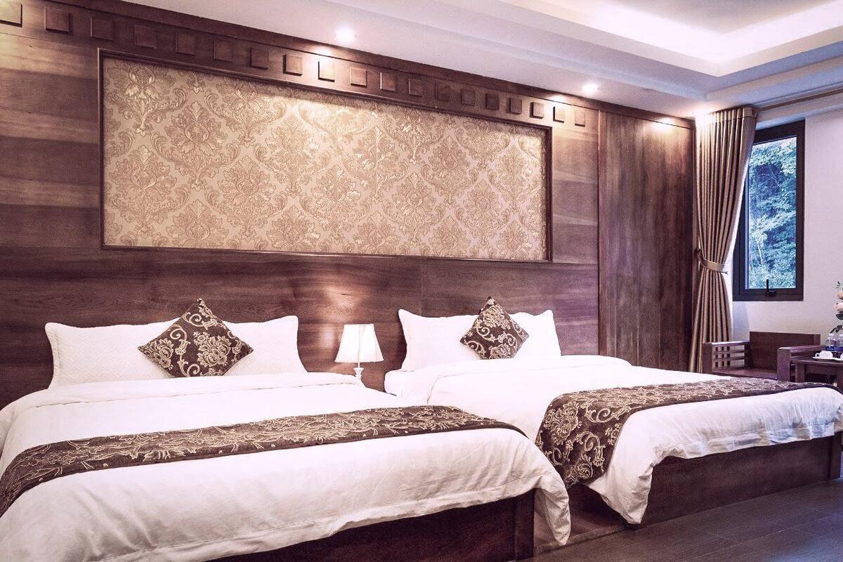 Quarto de hotel com duas camas, um painel decorativo grande, uma janela com cortinas e uma mesa pequena com abajur.