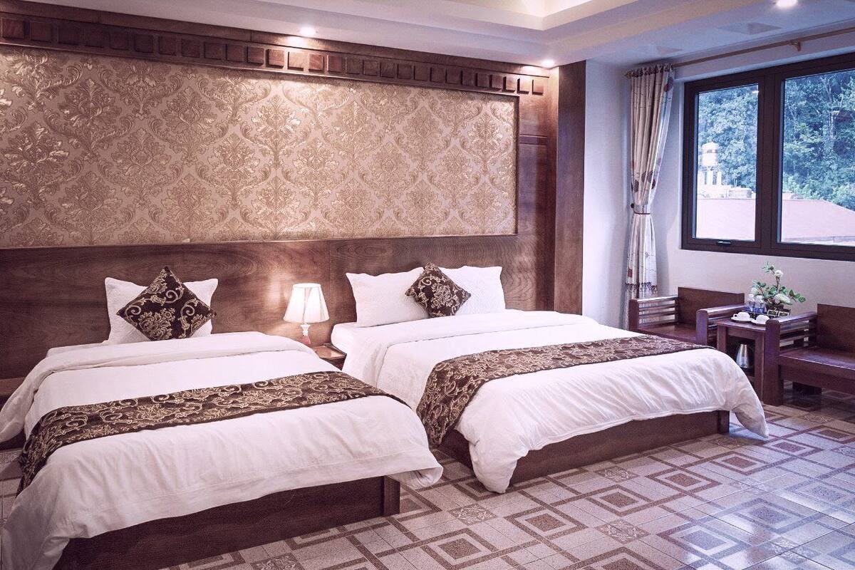 Quarto de hotel com duas camas, uma janela grande e uma cabeceira decorativa.