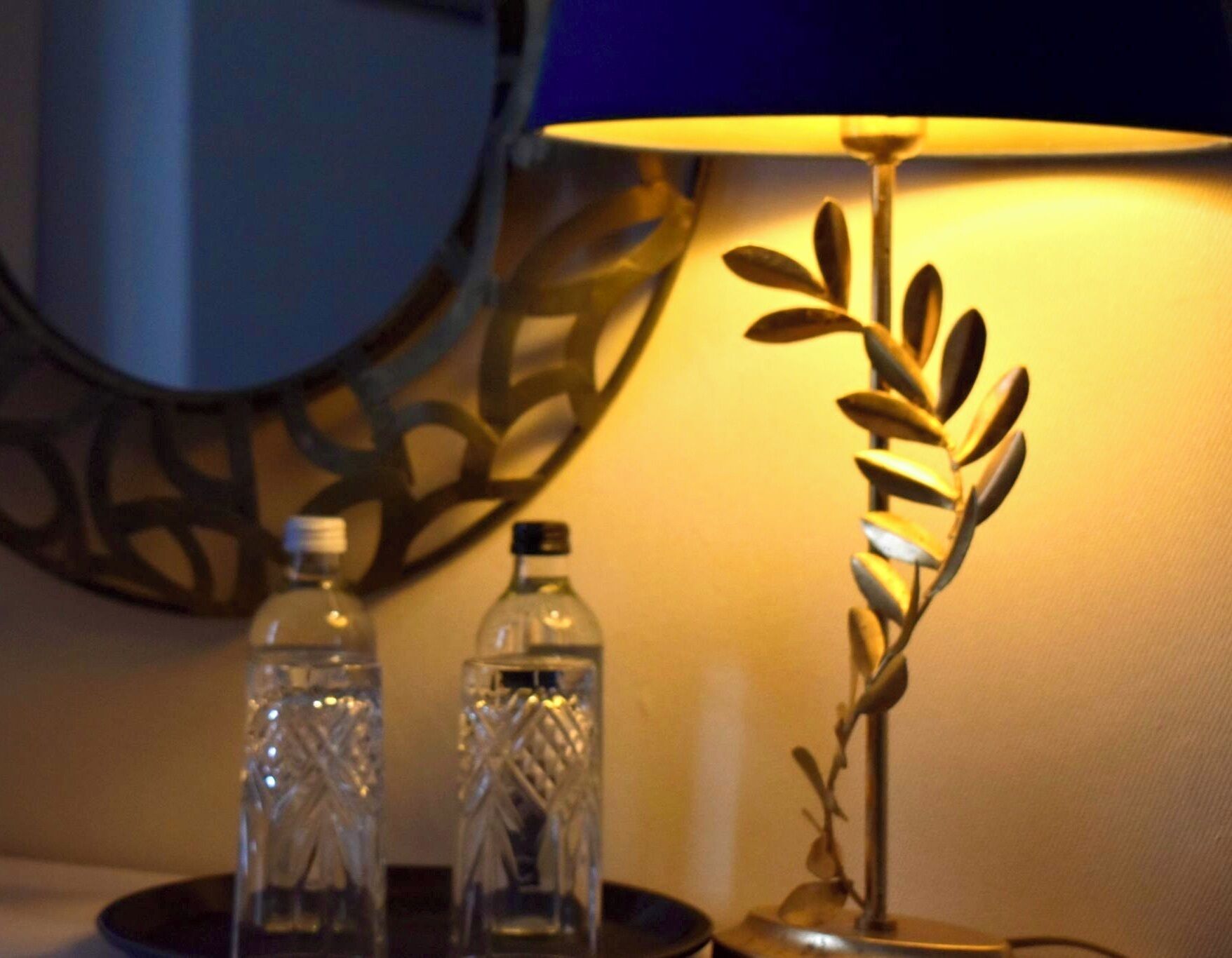 Uma mesa com duas garrafas de vidro transparente, um abajur de metal decorativo e um espelho fixado na parede.