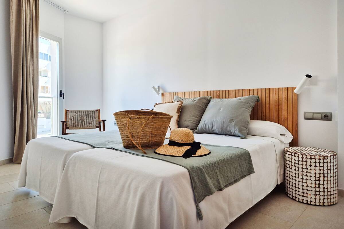 Uma cama bem arrumada com cabeceira de madeira, uma cesta de vime e um chapéu de palha.