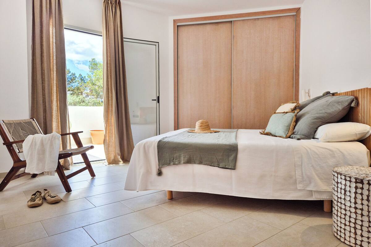 Quarto com cama, uma cadeira, uma mesinha e vista através de uma porta.