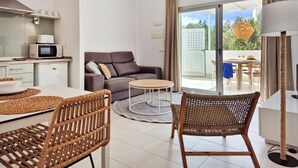 Sala de estar moderna com sofá, duas poltronas de vime, uma mesa de centro redonda e uma mesa de jantar com cadeiras. Há uma área de cozinha com micro-ondas e um refrigerador pequeno. O quarto possui uma grande porta de vidro deslizante que dá para um pátio externo com mesa e cadeiras.