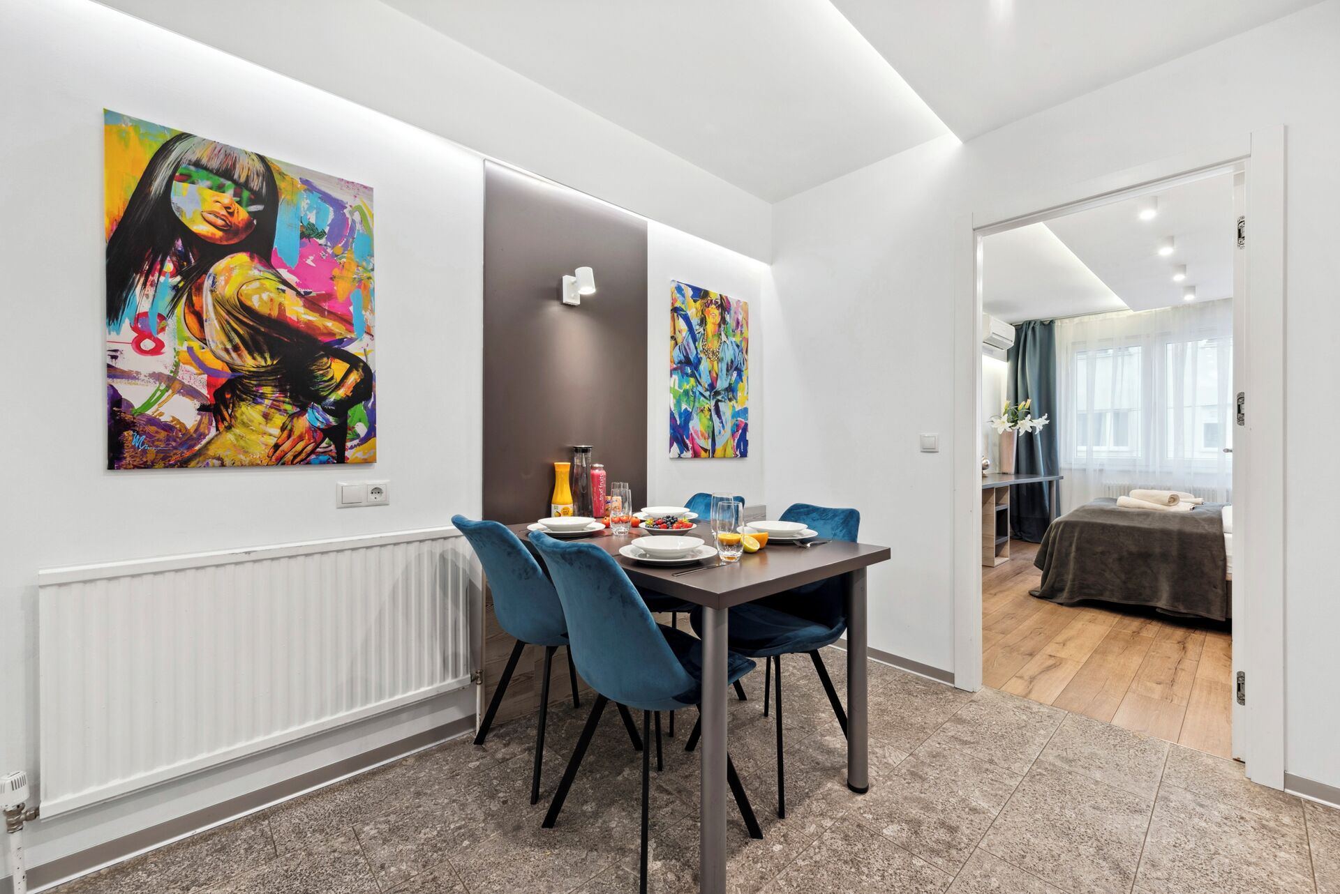 Uma sala de jantar moderna com uma mesa posta para dois, duas pinturas abstratas vibrantes nas paredes, um radiador e vista para um quarto com cama e cortinas.