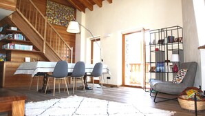 Uma sala de estar ampla com escada de madeira, área de jantar com mesa e cadeiras, estante de livros e um espaço de estar aconchegante com uma poltrona e um cesto.