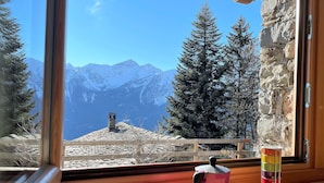 Vista pela janela: montanhas cobertas de neve, uma chaminé de pedra e uma cafeteira colorida sobre uma mesa de madeira.