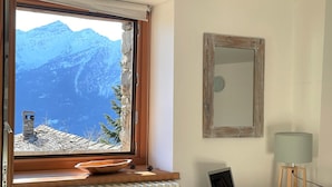 Quarto com vista para montanhas pela janela, uma escrivaninha de madeira com um laptop, um espelho e um abajur.