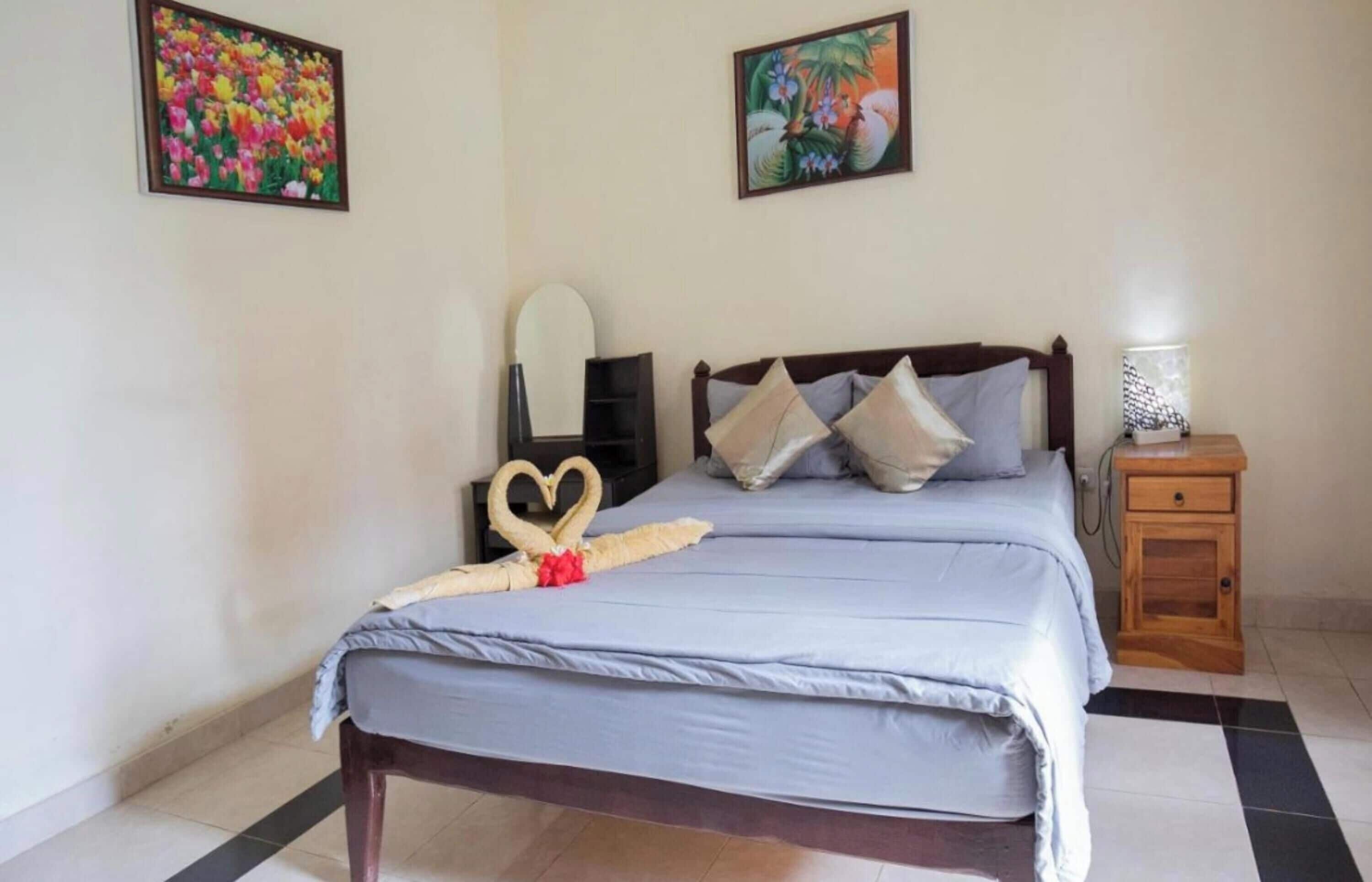 Quarto com cama, almofadas, mesa de cabeceira, abajur e duas pinturas fixadas na parede.