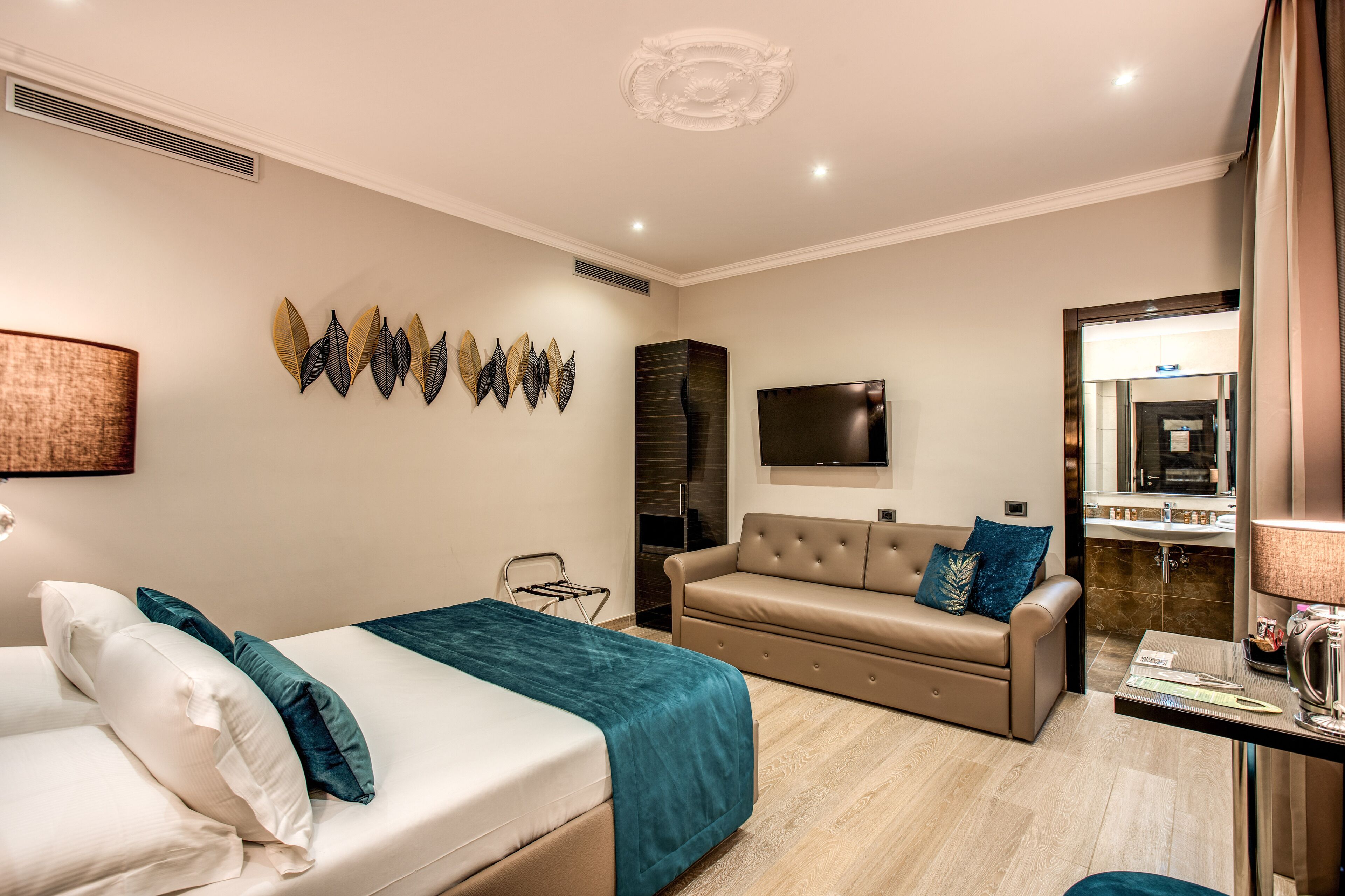 Quarto de hotel com cama, sofá, televisão, espelho e banheiro visível através de uma porta aberta.