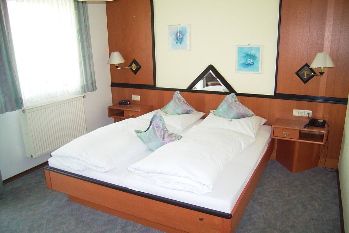 Uma cama de casal com duas almofadas, uma mesinha de cabeceira com abajur, um criado-mudo com telefone e uma janela com persianas.