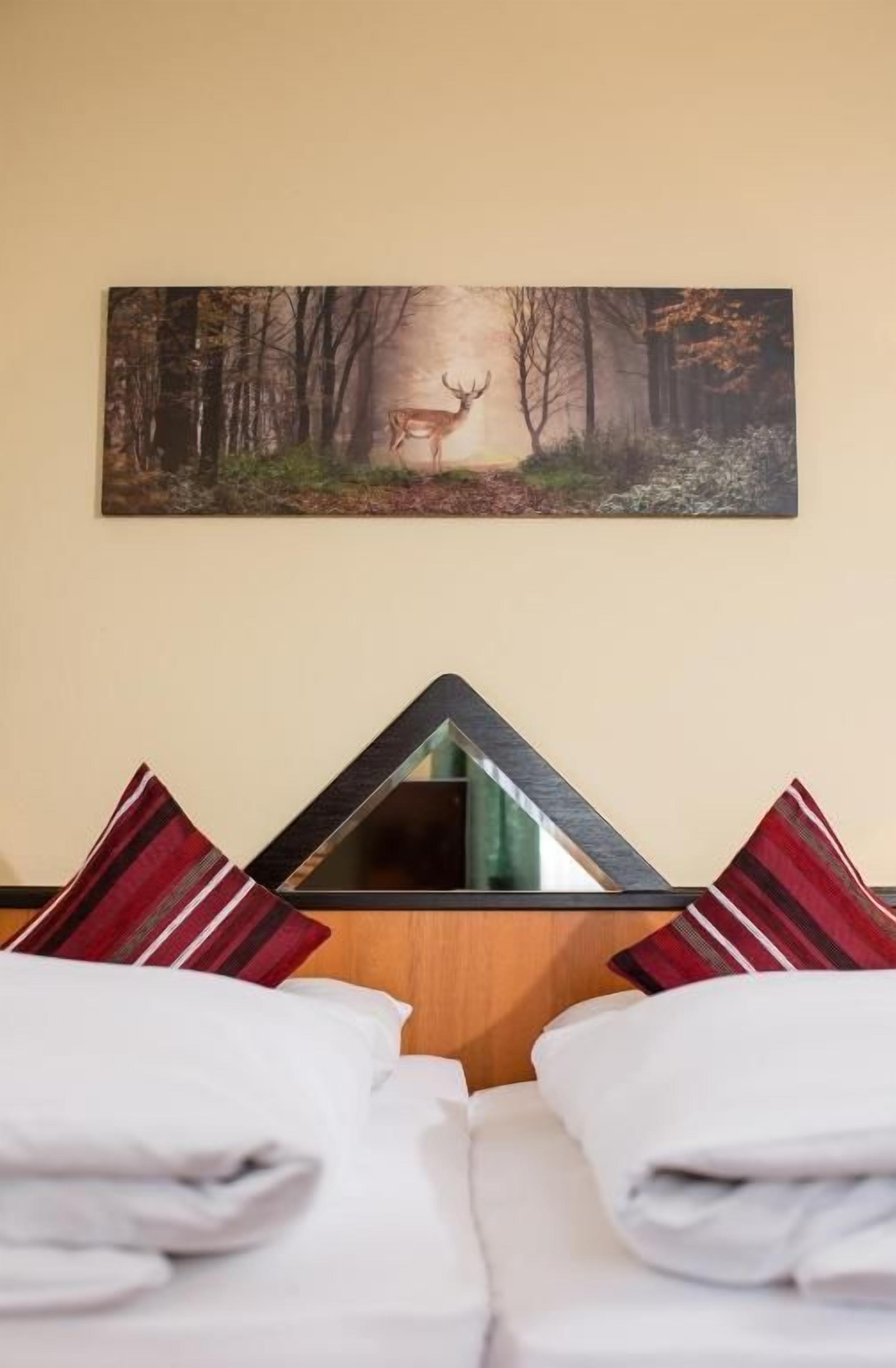 Quarto de hotel com uma cama, almofadas, um quadro de um veado numa floresta e um espelho na parede.