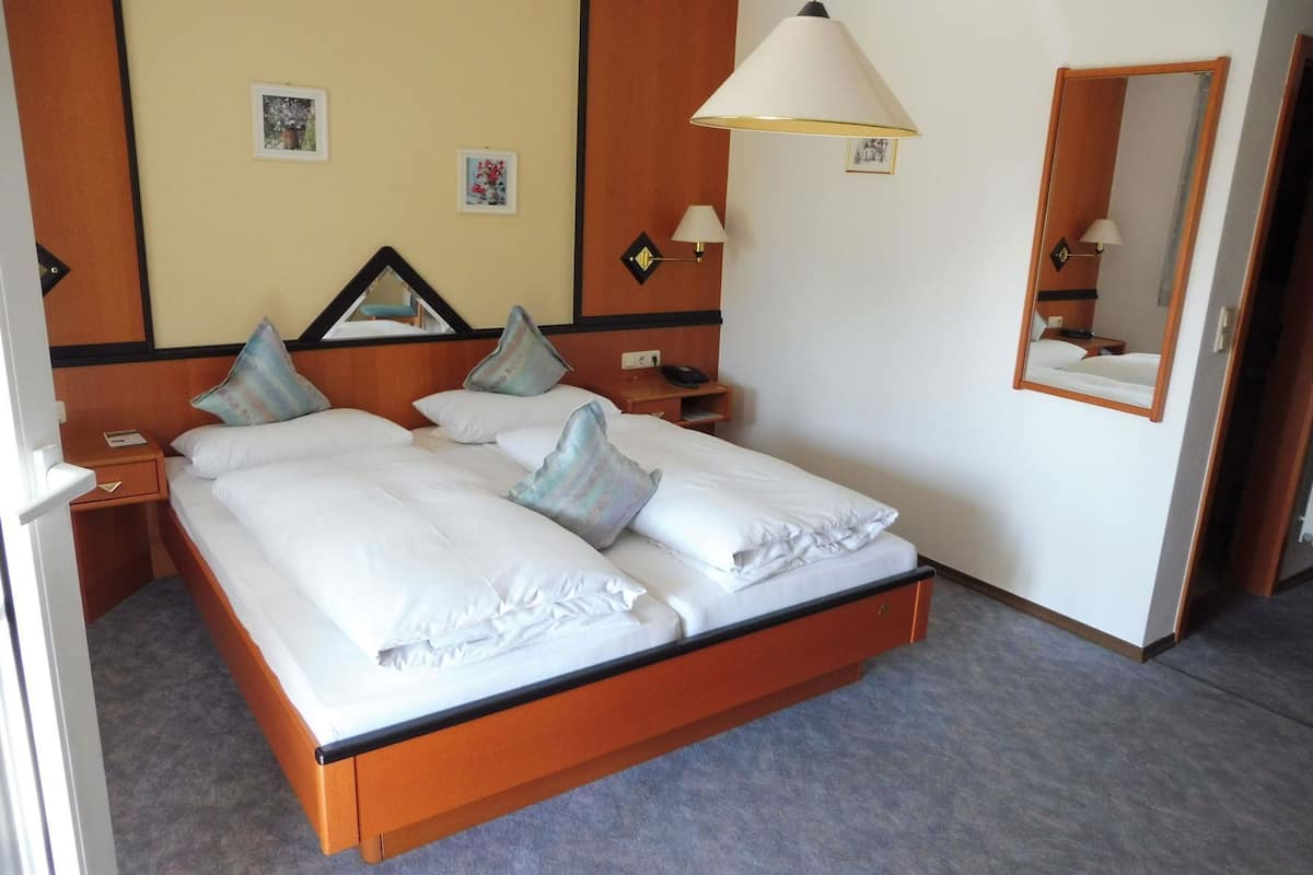 Uma cama de casal com lençóis brancos, uma mesa de cabeceira com abajur, um espelho e duas imagens emolduradas na parede.