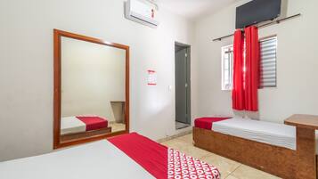 Quarto de hotel com um espelho grande, uma cama com colcha estampada em vermelho e branco, um banco de madeira com colchão branco, uma mesinha e uma televisão fixada na parede.