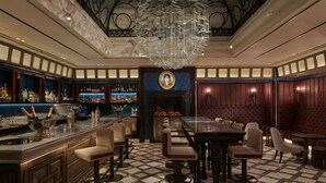 Uma área de bar com piso xadrez, banquetas, um grande candelabro e uma parede com um retrato emoldurado.