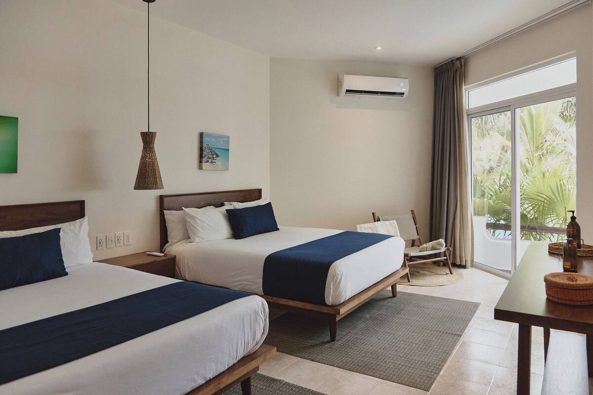 Quarto de hotel com duas camas, uma mesa, uma cadeira e uma janela grande com vista para palmeiras.