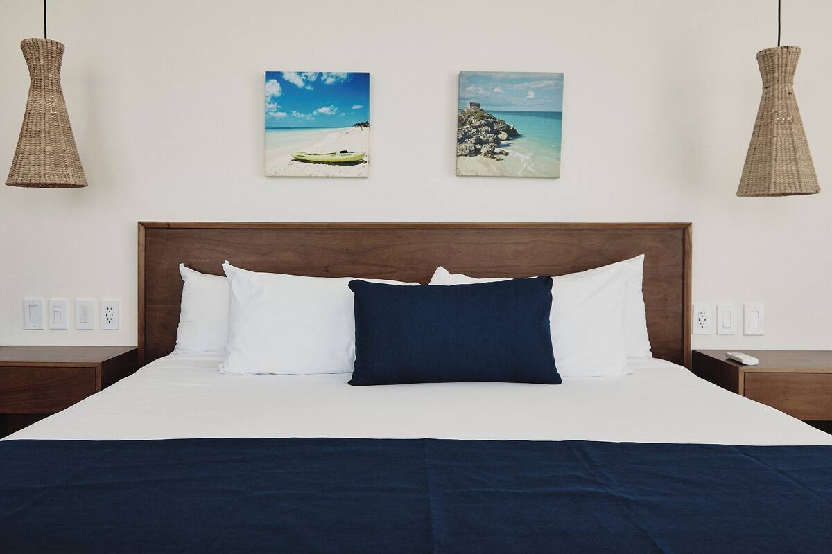 Uma cama bem arrumada com roupa de cama branca e azul escura, duas pinturas de cenas de praia penduradas na parede e duas luminárias pendentes de vime.