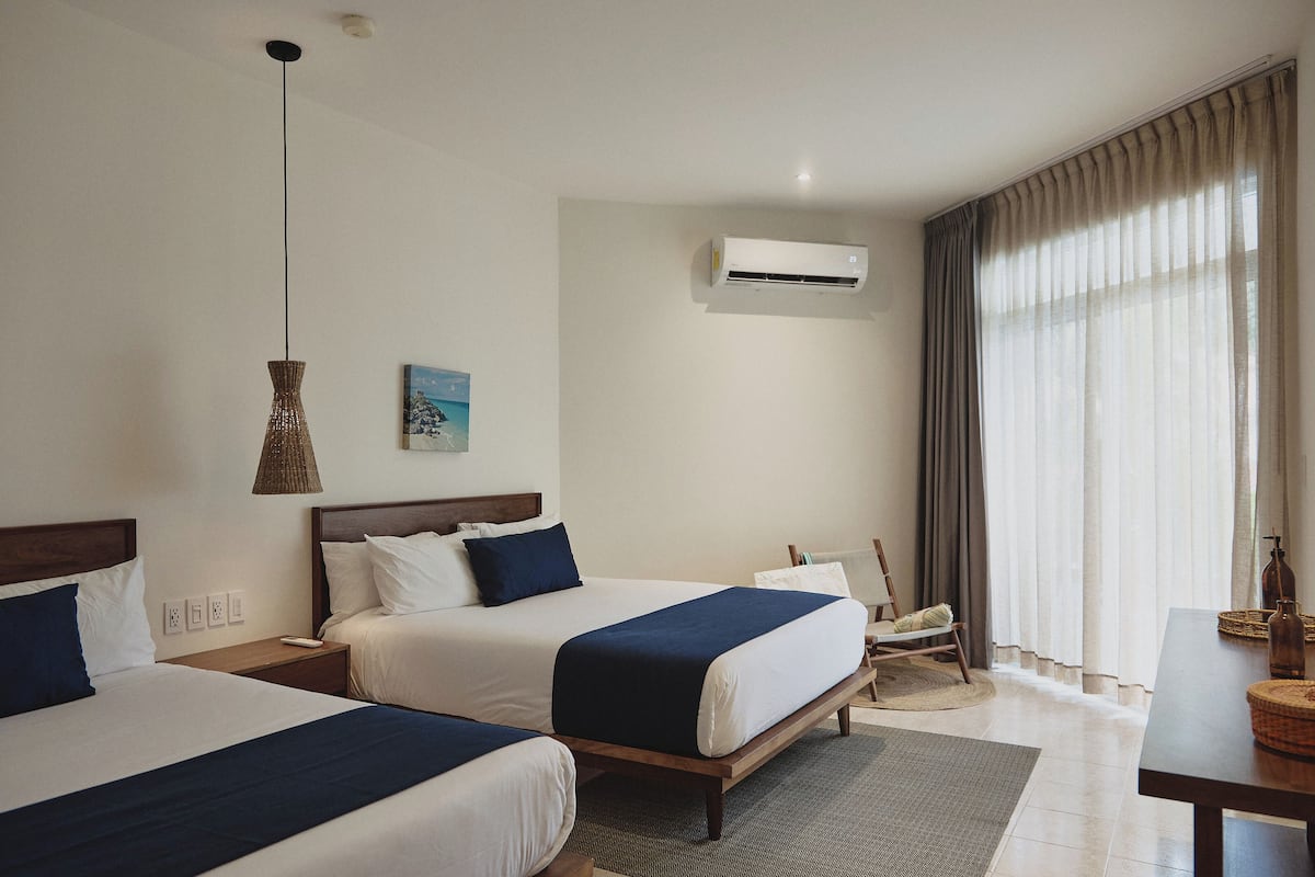 Quarto de hotel com duas camas, uma escrivaninha, uma cadeira, ar condicionado e uma janela grande com cortinas.