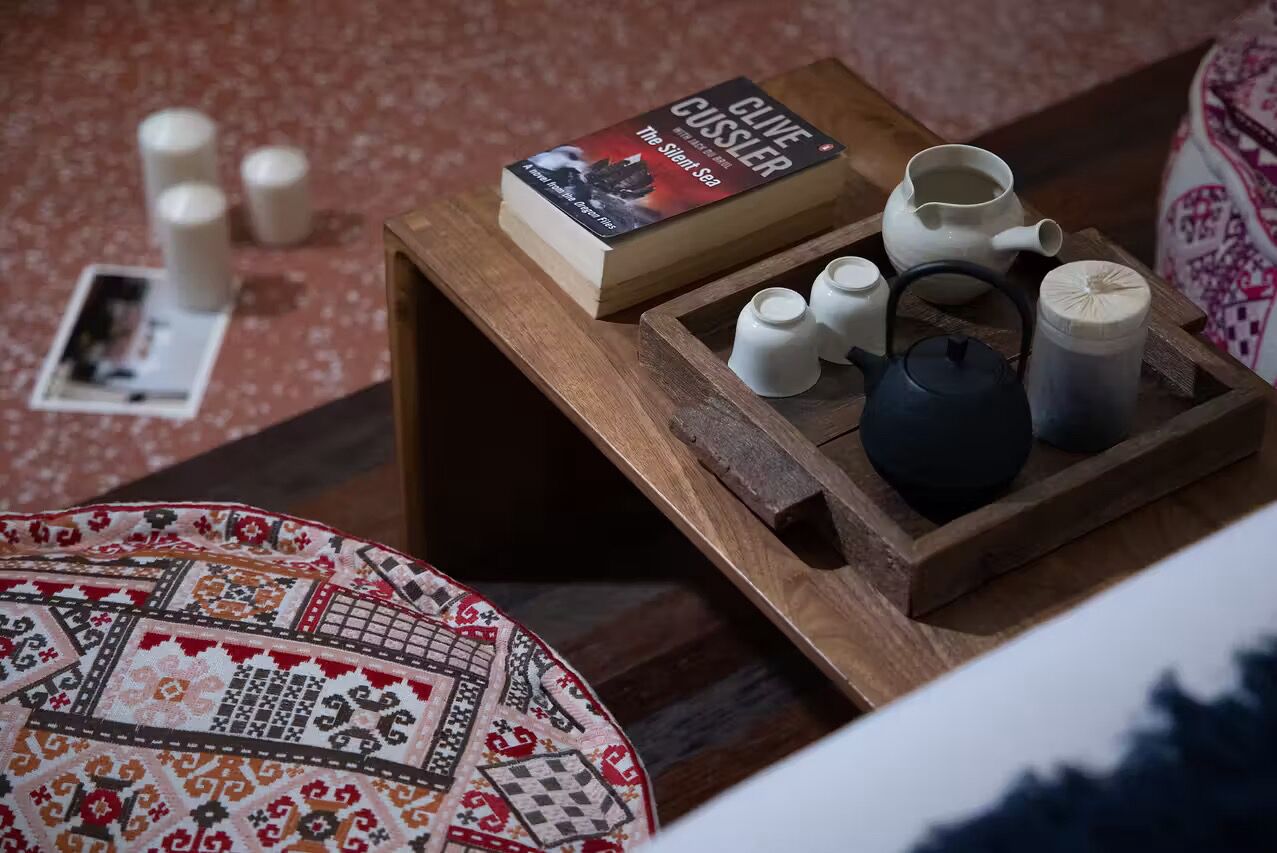 Uma mesinha de madeira com um livro intitulado 'The Silent Sea' de Clive Gussler, uma chaleira, xícaras e um tabuleiro.