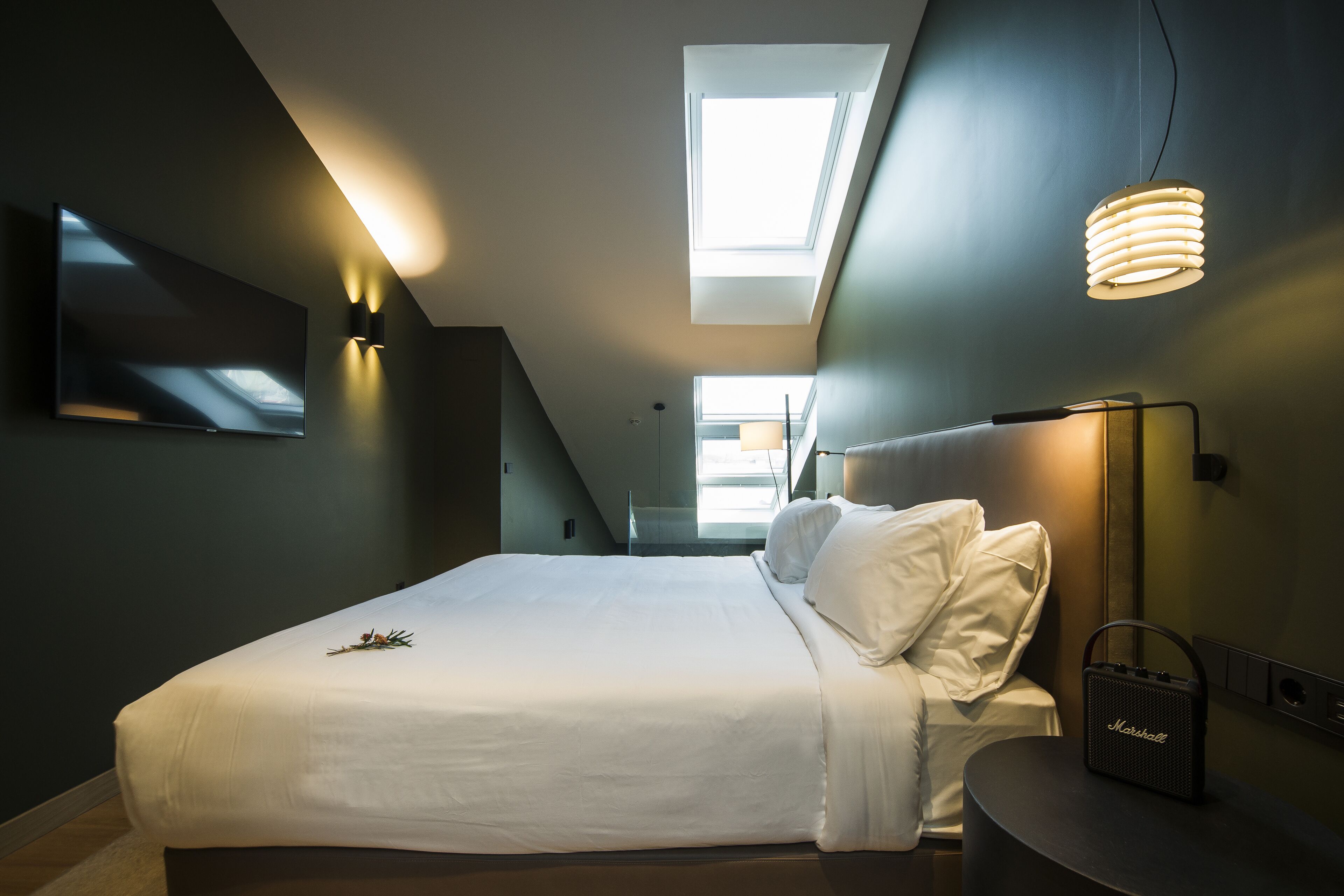 Quarto de hotel moderno com cama grande, TV de tela plana, claraboia e um alto-falante Marshall.