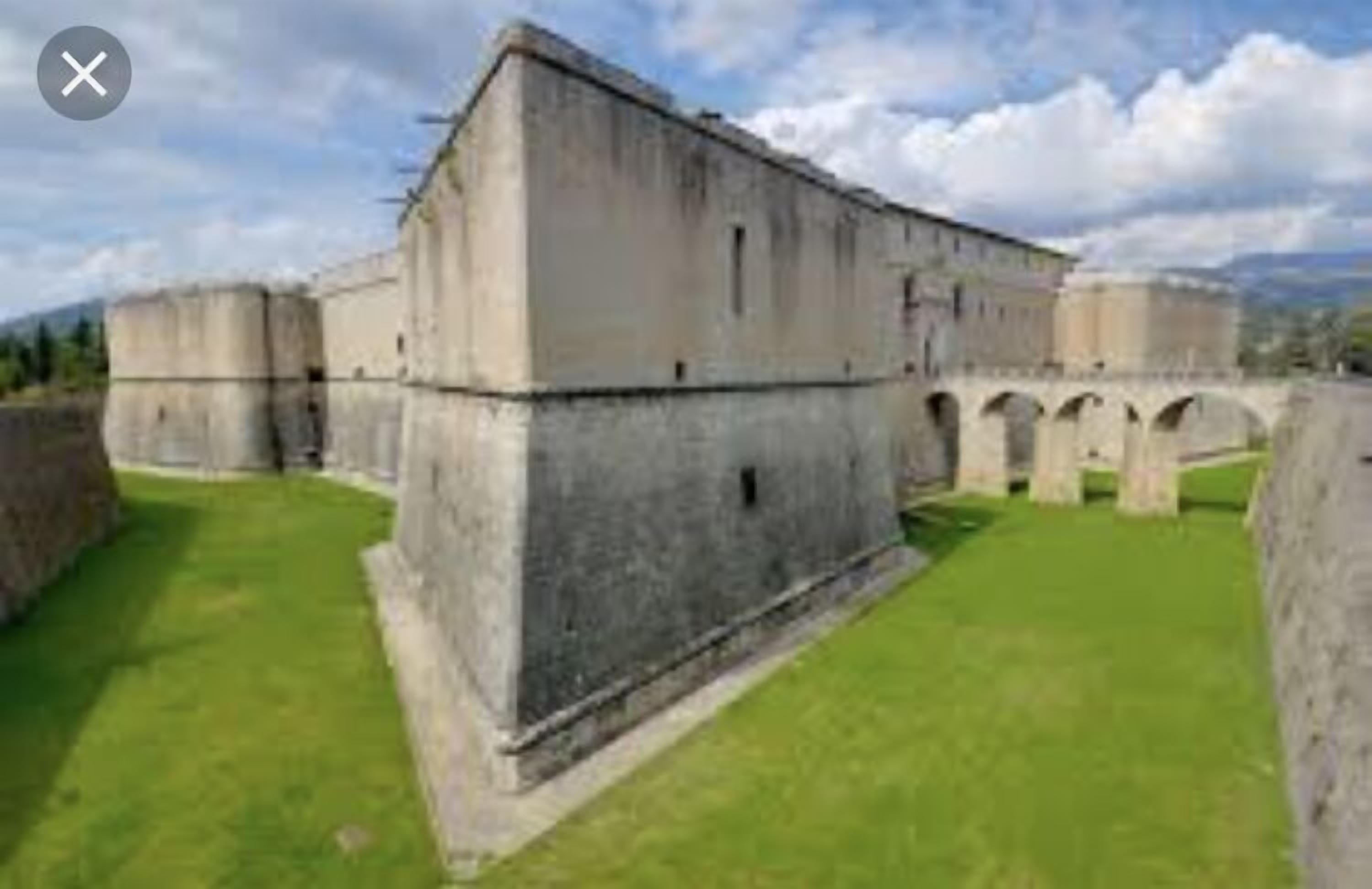 Um edifício grande e retangular com telhado plano, cercado por um muro de pedra baixo e um pátio gramado.