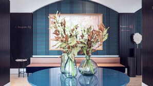 Uma área de recepção com uma mesa azul redonda, dois vasos de vidro com flores, um mapa na parede e um banco.